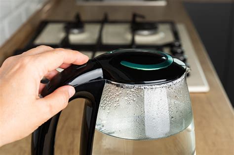 전기 주전자의 전원 스위치를 누르는 여성의 클로즈업 집에서 에너지를 절약하십시오 주방의 나무 테이블에 있는 현대적인 전기 투명 주전자 물을 끓이고 차를 만들기위한 주전자