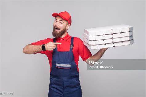 파란색 유니폼을 입고 수염과 젊은 행복 배달 남자와 빨간 T셔츠 서 들고 가리키는 회색 배경에 골 판지 피자 상자 더미를 손가락 유머에 대한 스톡 사진 및 기타 이미지