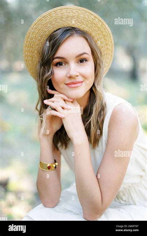 Young Woman In A Straw Hat Sits With Her Hands Clasped Under Her Chin