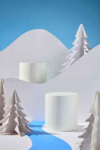 제품 전시를 위한 두 개의 빈 연단이 겨울 아이디어의 템플릿 이미지에 묘사되어 있으며 그 뒤에는 파란색 배경에 흰 종이 언덕이 있고 그 주변에는 소나무와 녹색 종이가