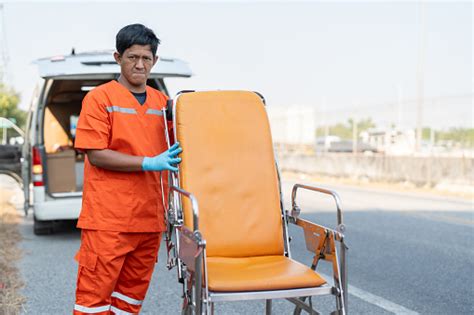 주황색 유니폼을 입은 선임 아시아계 남성 Ems 구급대원 또는 응급 의료 기술자가 길가의 구급차 근처 들것 옆에 서서 의료 응급 상황에서 조치를 취할 준비를 하고 있습니다