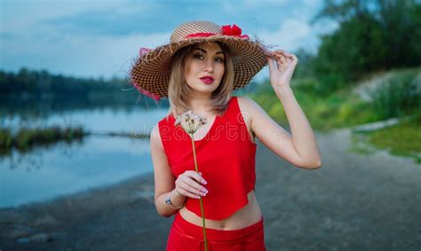 Девушка с цветком в красном платье и соломенной шляпе на речном береге в последнем вечере