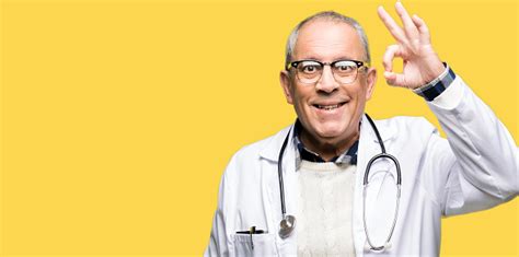 잘 생긴 의료 코트를 입고 잘생긴 수석 의사는 손과 손가락으로 확인 기호를 하 고 긍정적 웃는 성공적인 식입니다 건강관리와