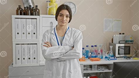 A Confident Young Hispanic Woman In A Lab Coat Poses In A Laboratory