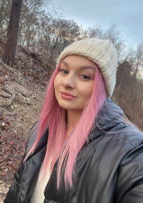 A Woman With Pink Hair Wearing A Black Jacket And White Hat Standing In