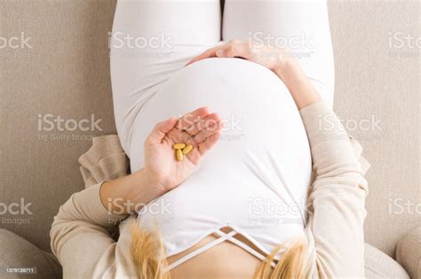 거실에서 소파에 앉아 큰 배를 가진 젊은 임산부 어머니와 미래의 신생아 건강을위한 손 들고 알약 임신 시간에 비타민을 받습니다