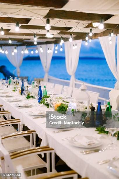 웨딩 디너 테이블 리셉션 바다에 대한 긴 테이블 하얀 식탁보 백열전구가 달린 화환 나무 흰색 의자입니다 테이블 중앙에 있는 꽃 조성물 결혼식에 대한 스톡 사진 및 기타