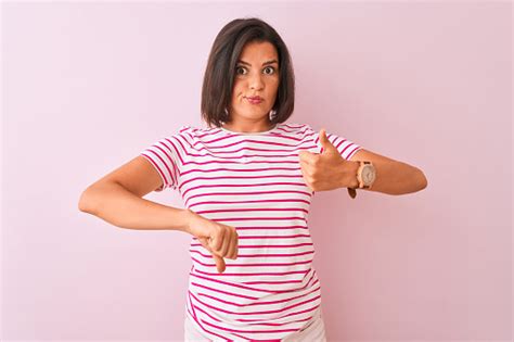 스트라이프 티셔츠를 입은 젊은 아름다운 여성은 고립 된 분홍색 배경 위에 서서 엄지 손가락을 위아래로 불일치와 동의 표현을하고 있습니다 미친 충돌 T 셔츠에 대한 스톡 사진