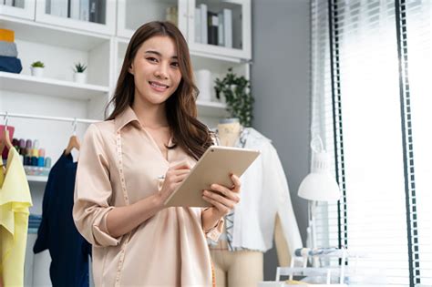 아틀리에를 재단사에서 옷을 입고 일하는 아시아 재단사 여성의 초상화 매력적인 아름다운 젊은 여성 패션 디자이너미소와 워크샵에서 새로운 수제 정장을 만들기에 성공 후 카메라를