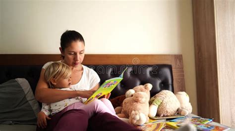 Mom Reads A Picture Book To Her Little Daughter Sitting On Her Lap On