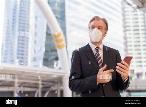 Mature Businessman With Mask Thinking While Using Phone At Skywalk