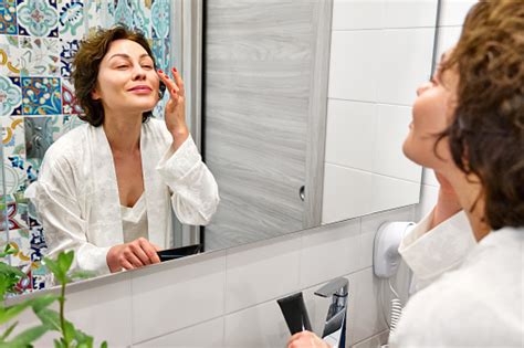 하얀 실크 목욕 가운을 입은 중년 여성이 욕실에서 거울을 보고 얼굴을 만지고 피부에 수분 공급 안티에이징 화장품을 바르고 웃고