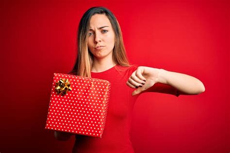 화가 얼굴 엄지 손가락으로 싫어하는 것을 보여주는 부정적인 기호 거부 개념과 빨간 배경 위에 생일 선물을 들고 푸른 눈을 가진 젊은 아름다운 금발의 여자 개념에 대한 스톡