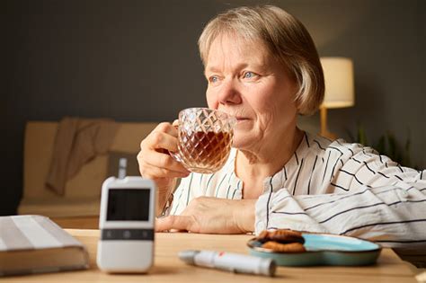 나이 든 여자가 나무 테이블에 앉아 유리잔에 담긴 차를 홀짝이고 있다 그녀는 따뜻하고 매력적인 방에서 쿠키와 공책을 들고 주변을 관찰합니다 55 59세에 대한 스톡 사진 및