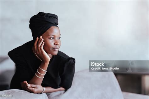 검은 터번과 전통 드레스를 입은 잠겨 있는 아프리카 소녀는 빈 공간이 있는 벽에 소파에 기대어 옆으로 보입니다 사려 깊은 젊은 성인 여성이 집에서 휴식을 취하고 있습니다