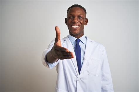 고립 된 흰색 배경 위에 서있는 코트를 입은 젊은 아프리카 계 미국인 의사가 인사와 환영으로 악수를 제공합니다 성공적인 비즈니스 몸짓에 대한 스톡 사진 및 기타 이미지