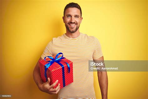 행복한 얼굴서서 이빨을 보여주는 자신감 있는 미소로 미소 짓는 노란 고립된 배경 위에 생일 선물을 들고 있는 청년 건강한 생활