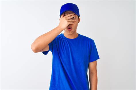 파란색 티셔츠와 모자를 쓰고 고립된 흰색 배경 위에 서있는 중국인 배달원이 얼굴과 눈을 손으로 덮고 당황한 표정으로 손가락을