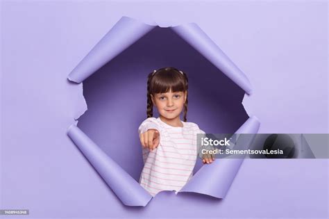 줄무늬 셔츠를 입은 피그 테일을 가진 진지한 어린 소녀의 초상화는 보라색 종이 배경을 통해 깨지고 여성 아이가 손가락과 카메라를 가리키고 당신을 선택하고 그녀의 선택을 보여