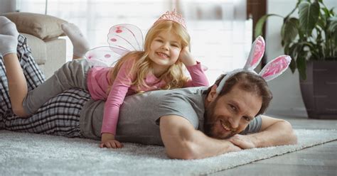 재미있는 게임 즐거운 남자의 초상화는 아이를 등에 업고 바닥에 누워 있다 그는 카메라를 바라보며 웃고 있다 소녀는 요정 날개와 분홍색 왕관으로 포즈를 취하고 있다 사진
