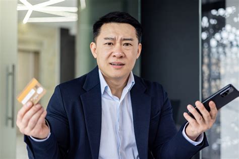 사무실 내부의 실망하고 화난 사업가의 초상화 은행 신용 카드와 전화를 들고 카메라를 화나게하는 속임수 아시아 남자 온라인