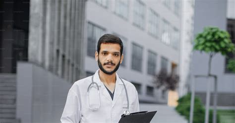 흰색 코트를 입은 젊은 잘생긴 수염을 기른 아라비아 남자 의사 의료 종사자의 전체 길이 초상화 현대 병원이나 클리닉 앞 야외에서 클립보드 폴더를 손에 들고 서 있습니다