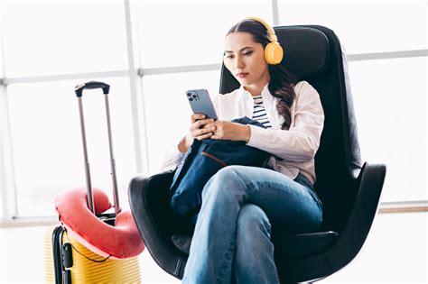 공항 라운지에서 헤드폰을 끼고 여행 가방을 옆에 두고 스마트폰을 확인하고 있는 매력적인 여성 20 24세에 대한 스톡 사진 및 기타 이미지 Istock