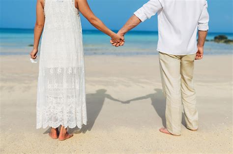 흰 옷을 입은 남자와 여자가 뒤로 서서 손을 잡고 바다에서 결혼식 전 사진 촬영을합니다 땅에 그림자가 드리워져 있었다 미래의 신부와 신랑으로서 결혼을 준비합니다 신랑 결혼식