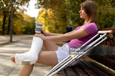 Woman With A Cast On Her Leg Sitting On A Bench In A Park And Drinking