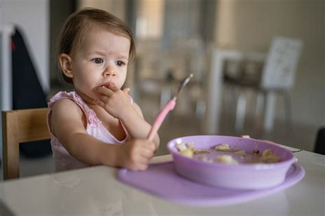 한 아이 작은 백인 유아 여성 아기는 방에서 집에서 혼자 테이블에서 먹는 숟가락을 사용하여 실제 사람들이 공간을 복사합니다 조기 아동 발달 개념 학습 프론트 뷰는 음식을