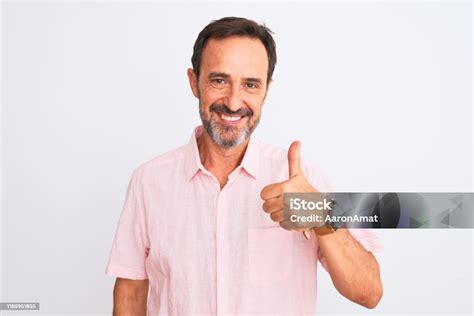 고립 된 흰색 배경 위에 서있는 캐주얼 한 분홍색 셔츠를 입은 중년 잘생긴 남자가 행복한 엄지 손가락으로 손을 들고 제스처를 취합니다 성공을 보여주는 카메라를 보고 표정을