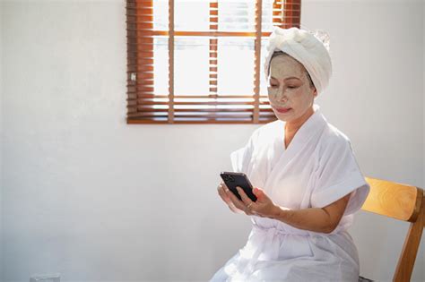 아시아 노인 여자는 얼굴 피부 건강을 위해 나무 의자에 흰색 코트 독서 노트와 휴대 전화를 착용 하 여 얼굴 크림을 적용