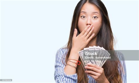 젊은 아시아 여 자가 고립 된 배경 표지 입 손으로 이상의 달러를 들고 실수 두려움 침묵 비밀 개념에에서 무서 워의 표현에 대 한 수치와 충격 가리기에 대한 스톡 사진 및