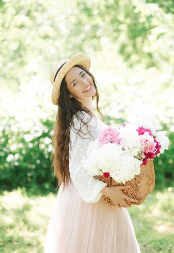 하얀 분홍색과 분홍색 모란 꽃이 든 고리 버들 바구니를 들고 밀짚 모자를 쓰고 유럽 도시의 거리에서 포즈를 취하는 행복한 미소