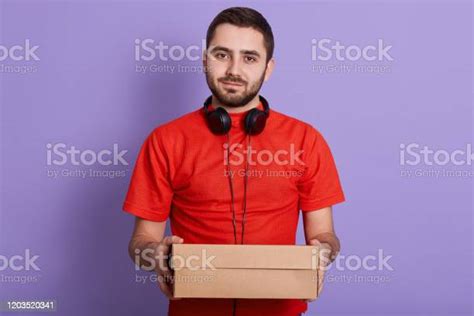 스튜디오에서 라일락 배경 위에 고립 된 젊은 수염 배달 남자의 절반 길이 초상화 양손에 카드 박스를 들고 즐거운 얼굴 표정을 가지고 목 주위에 헤드폰을 가지고 가득 찬에