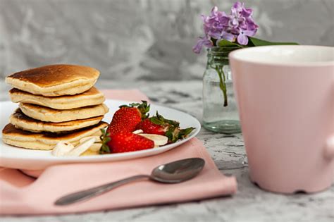 밝은 배경에 흰색 접시에 분홍색 차 찻잔과 신선한 딸기가있는 팬케이크 더미 아침 식사 옆에는 유리 꽃병에 담긴 작은 라일락 꽃다발이 있습니다 건강한 생활방식에 대한 스톡