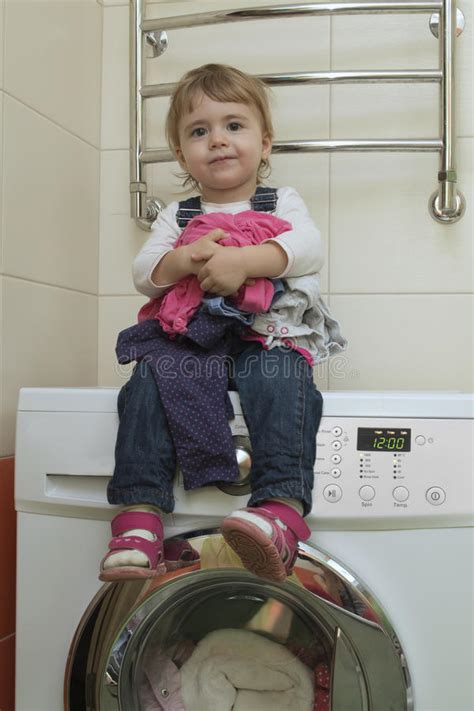 Счастливая милая маленькая девочка при одежды сидя на стиральной машине в домашнем интерьере