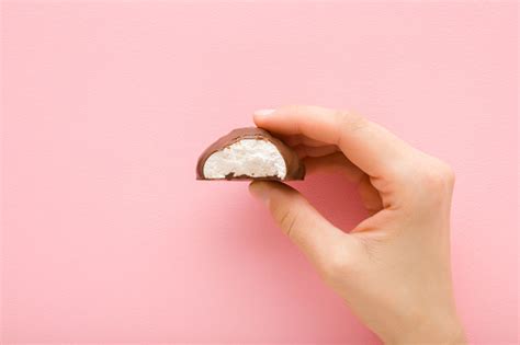 밝은 분홍색 테이블 배경에 짙은 갈색 초콜릿 유약으로 덮인 물린 흰색 바닐라 부드러운 제퍼를 들고 있는 젊은 성인 여성 파스텔 색상 근접 촬영 달콤한 간식 하향식 보기