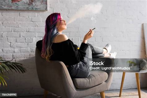 時髦的女孩與五顏六色的頭髮坐在扶手椅和吹煙的側視圖 照片檔及更多 吸煙 照片 吸煙 大麻 大麻草 大麻香煙 Istock