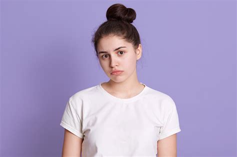 라일락 벽에 포즈를 취하는 슬픈 표정으로 어두운 머리의 아가씨가 카메라를 바라보며 기분이 좋지 않고 흰색 티셔츠를 입고 화가 난
