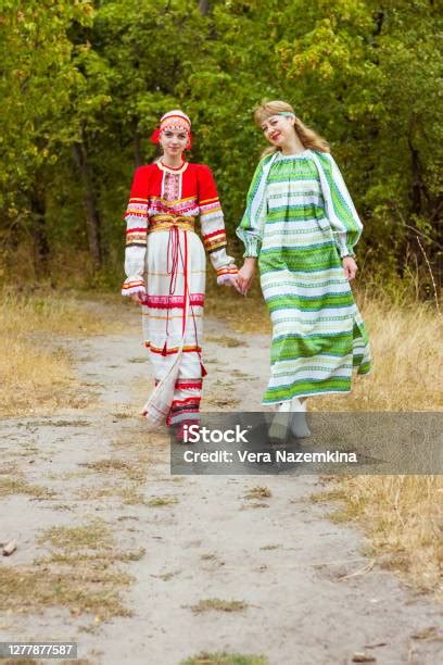 빨간색과 녹색의 민속 의상을 입은 서로 다른 연령대의 두 젊은 여성이 손을 잡고 공원의 길을 따라 걷습니다 14 15 살에 대한