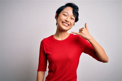 고립 된 흰색 배경 위에 서있는 캐주얼 한 빨간 티셔츠를 입은 젊은 아름다운 아시아 소녀는 전화통화처럼 손과 손가락으로 전화