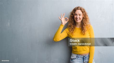 그런 지 회색 벽 손과 손가락으로 Ok 사인을 하 고 긍정적인 미소를 통해 젊은 빨강 머리 여자 성공적인 식입니다 곱슬 머리에