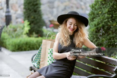 검은 펠트 모자와 쇼핑백드레스에 좋은 젊은 여성은 도시 공원의 벤치에 앉아 스마트 폰을 사용 30 34세에 대한 스톡 사진 및