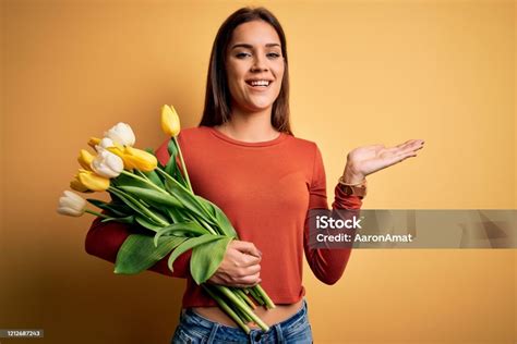 젊은 아름 다운 갈색 머리 여자 노란 배경 에 튤립 꽃다발을 들고 쾌활 한 제시 하 고 카메라를 보고 손바닥으로 가리키는 미소