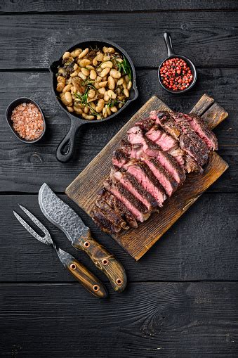 슬라이스 구운 고기 스테이크 립 눈 중간 희귀 나무 서빙 보드에 주철 팬에 흰색 콩과 로즈마리 고기 칼과 포크 검은 나무 테이블