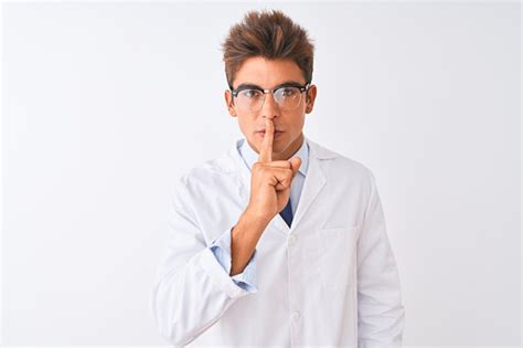 고립 된 흰색 배경 위에 안경과 코트를 입은 젊은 잘생긴 Sciencist 남자는 입술에 손가락으로 조용히 하도록 요청합니다