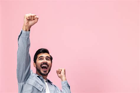 턱수염 행복 승리를 가진 갈색 머리 남자의 패션 초상화는 흰 티셔츠 미소와 그의 얼굴에 기쁨의 감정에 분홍색 배경에 주먹을 들고 손을 들어 올렸다 복사 공간 25 29세에