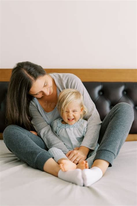 Smiling Mom Hugs A Little Laughing Girl Sitting Behind Her On The Bed