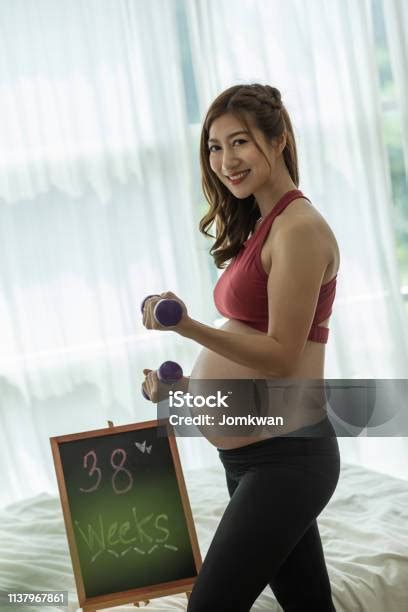 행복 한 아름 다운 매력적인 임신한 아시아 여자 창 및 운동에 가까이 서 있는 아령 미소와 함께 운동 하는 큰 배꼽 느낌 그래서